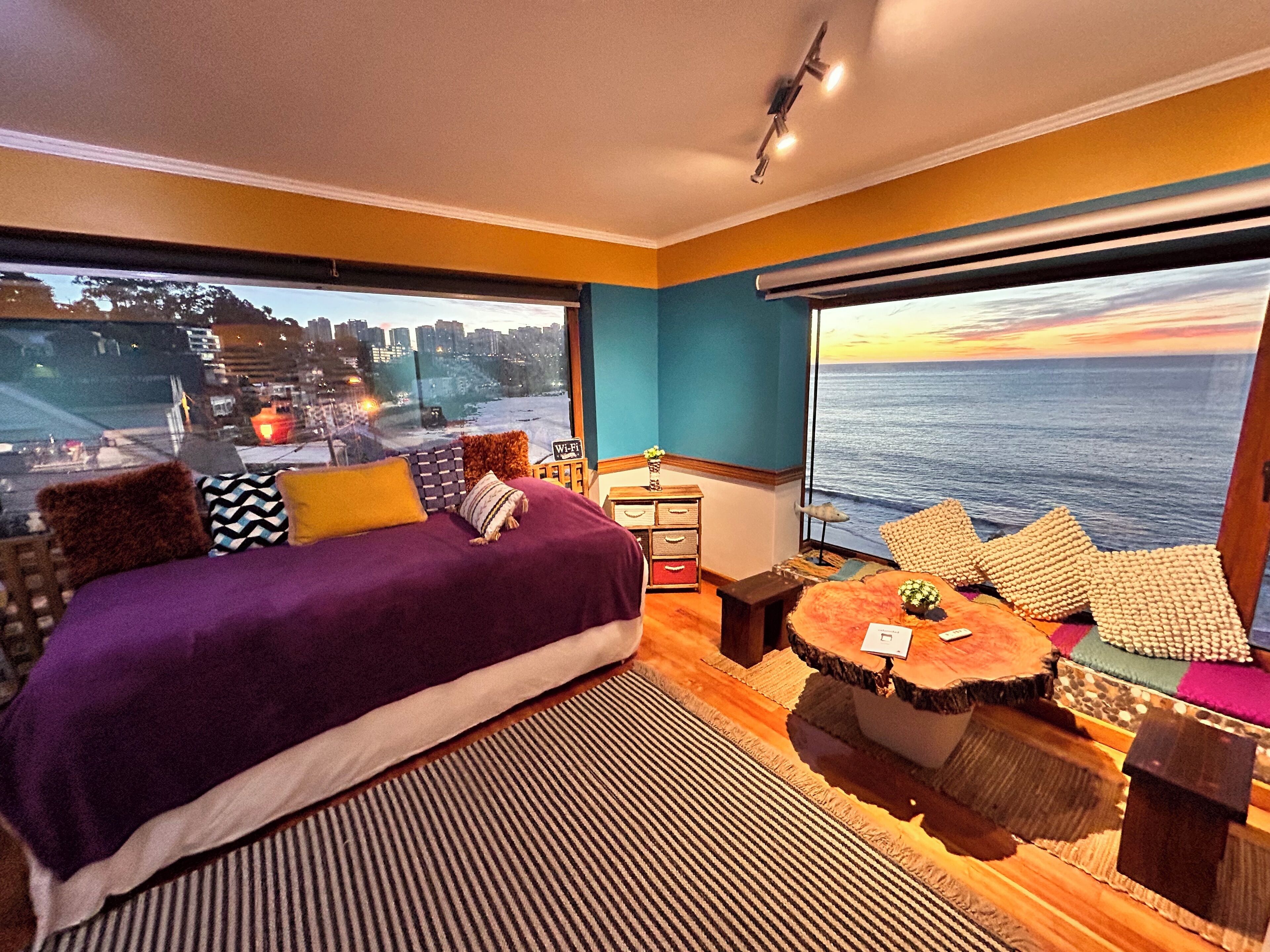 Quarto moderno com uma cama grande, um sofá pequeno, uma mesa de centro e vista para a cidade e oceano.