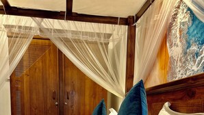 Cama com lençóis brancos e almofadas azuis, guarda-roupa de madeira e cama de dossel com cortinas leves.