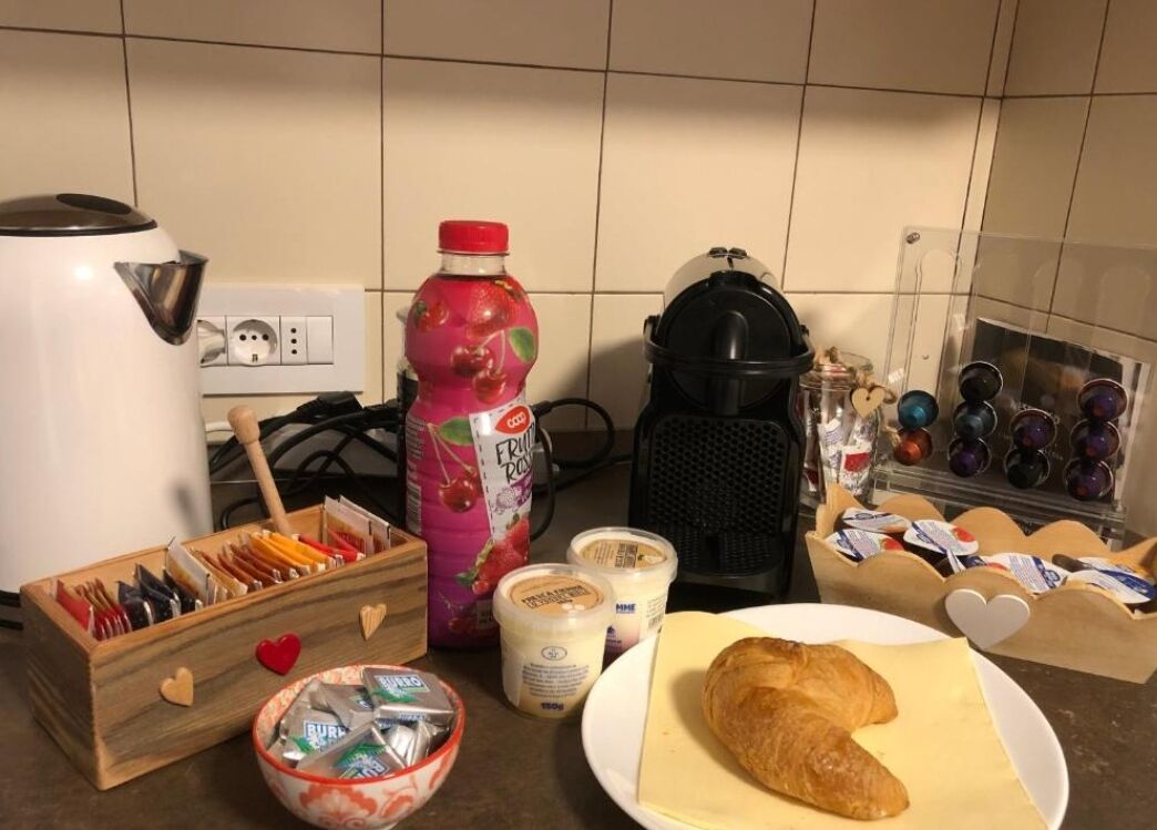 Uma bancada de cozinha com uma cafeteira, uma garrafa de ponche de frutas, um moedor de café preto, um recipiente com lanches, um prato com um croissant e um pote de iogurte.