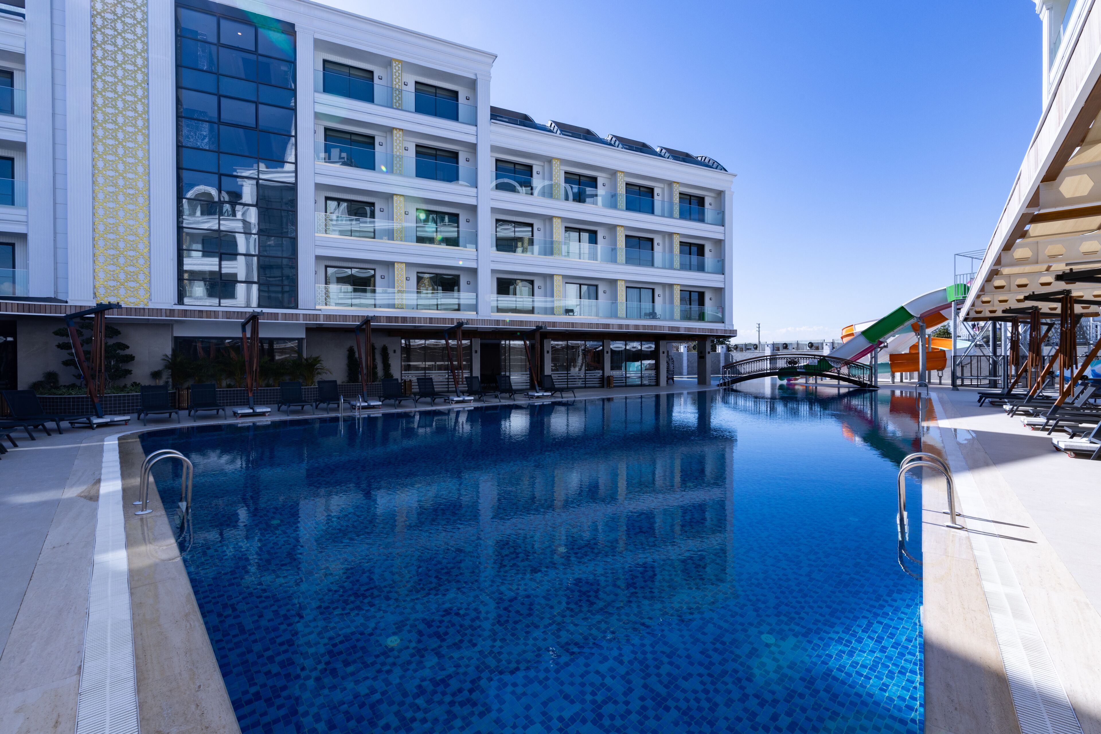 Piscina com superfície de água azul cristalina, rodeada por espreguiçadeiras e um edifício moderno com várias varandas.