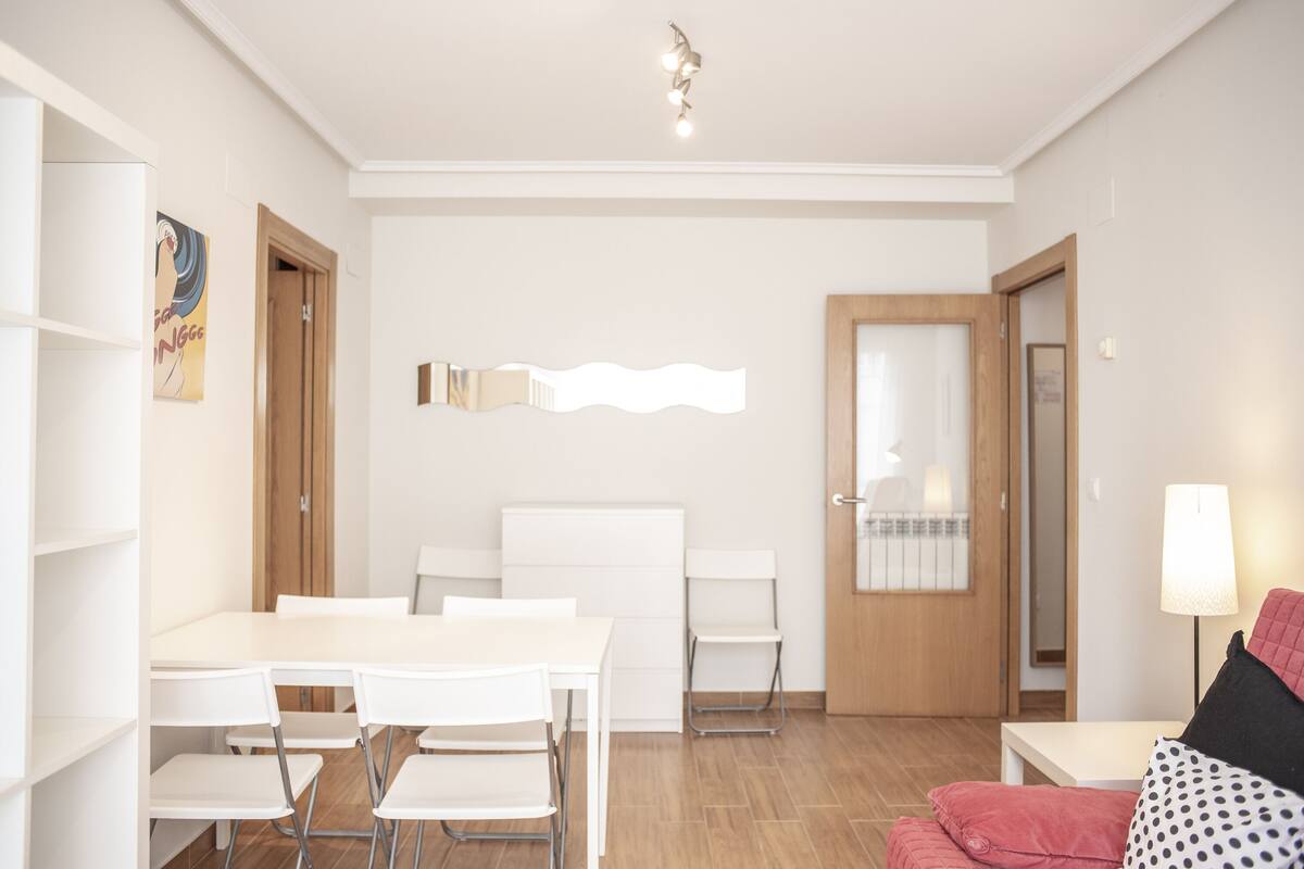 Quarto moderno e bem iluminado com mesa de jantar, cadeiras, sofá e abajur.