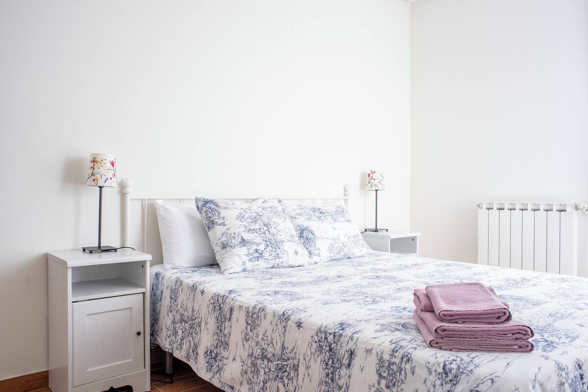 Cama bem arrumada com colcha floral, luminária de cabeceira e toalha dobrada no criado-mudo.