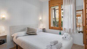 Cama bem arrumada com lençóis brancos, almofada listrada e toalhas enroladas ao lado. Há uma janela com moldura de madeira e cortinas brancas, e uma pequena mesa de cabeceira com uma luminária.