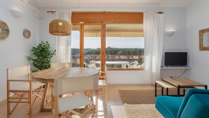 Uma sala de estar moderna com uma mesa de jantar de madeira, cadeiras, um sofá, uma TV e uma janela grande com vista para a cidade.