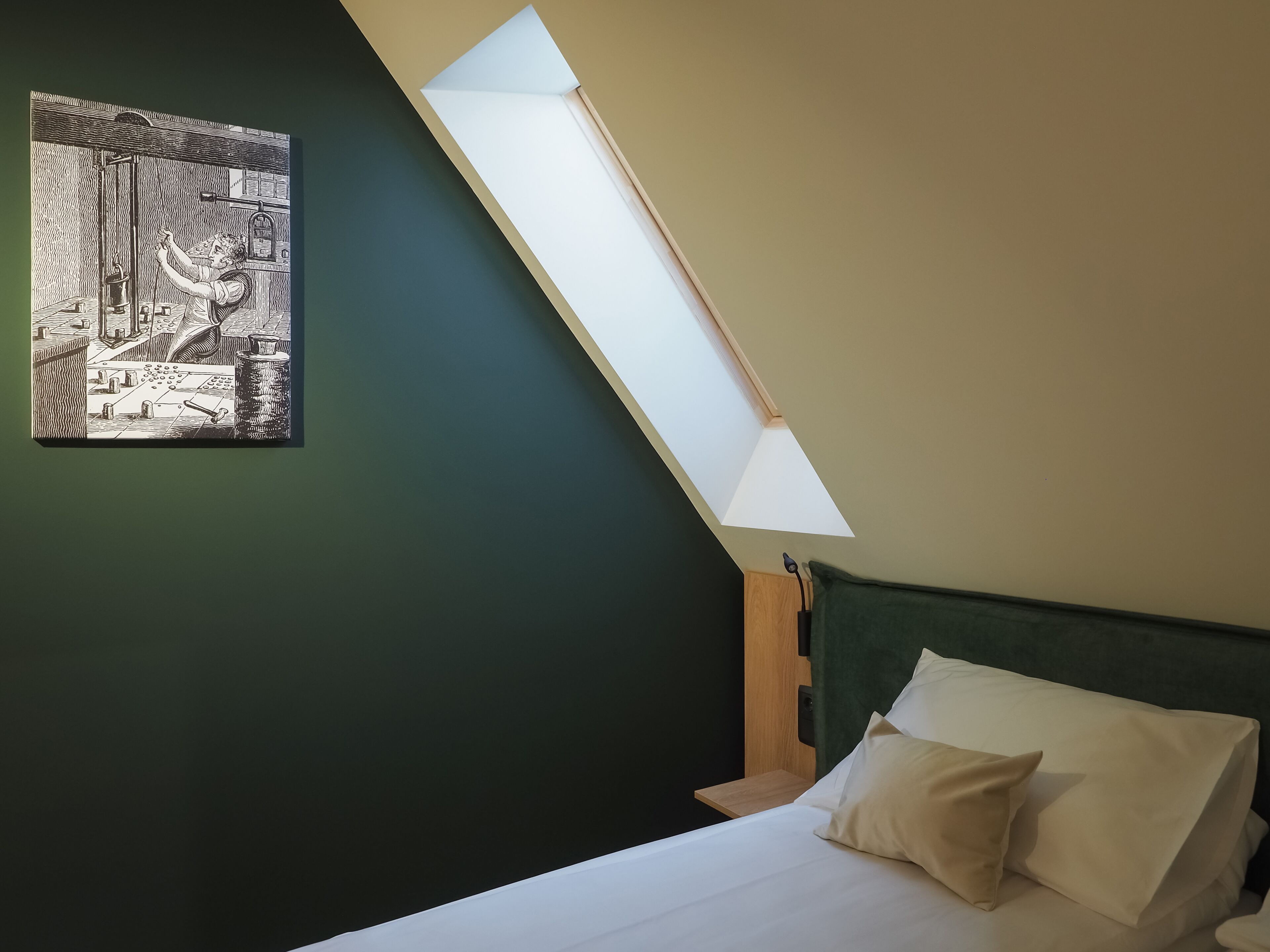 Quarto com claraboia, cama com roupa de cama branca, parede verde e uma obra de arte emoldurada.