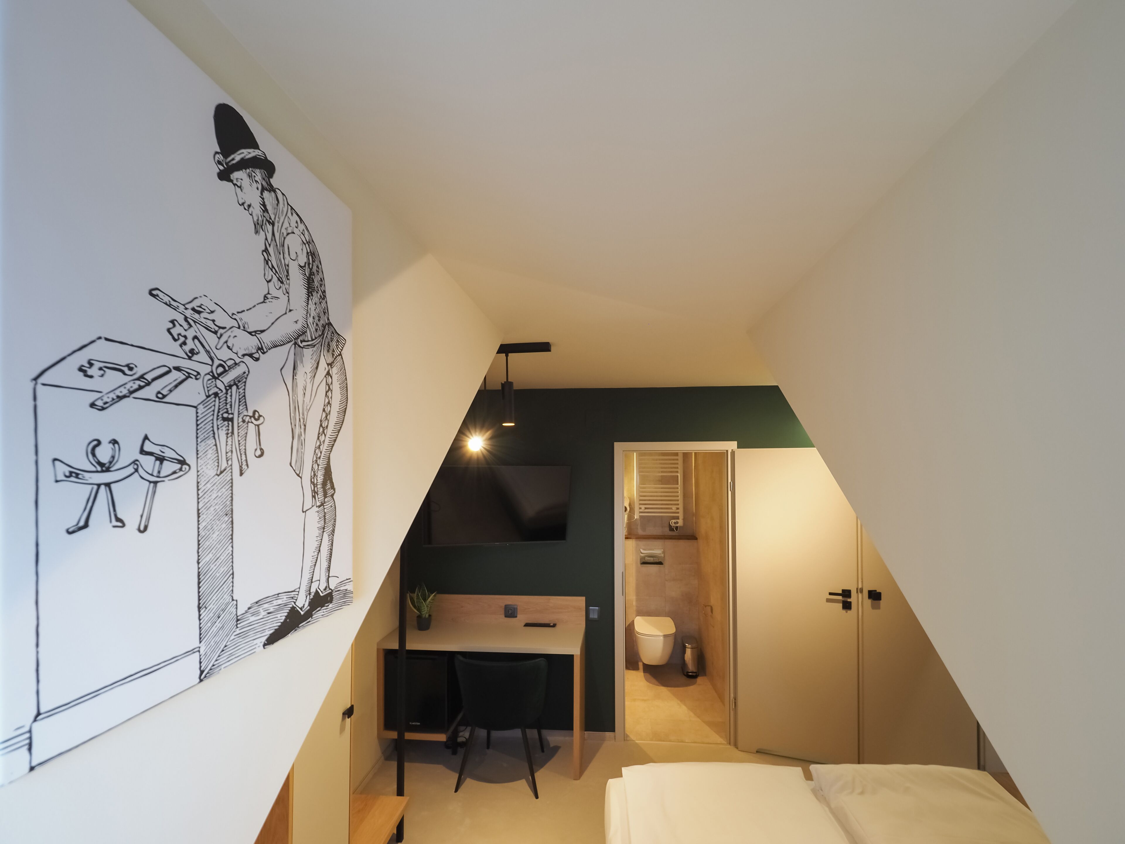 Um quarto pequeno e moderno, com escrivaninha e cadeira, uma cama e um banheiro visível através de uma porta aberta.