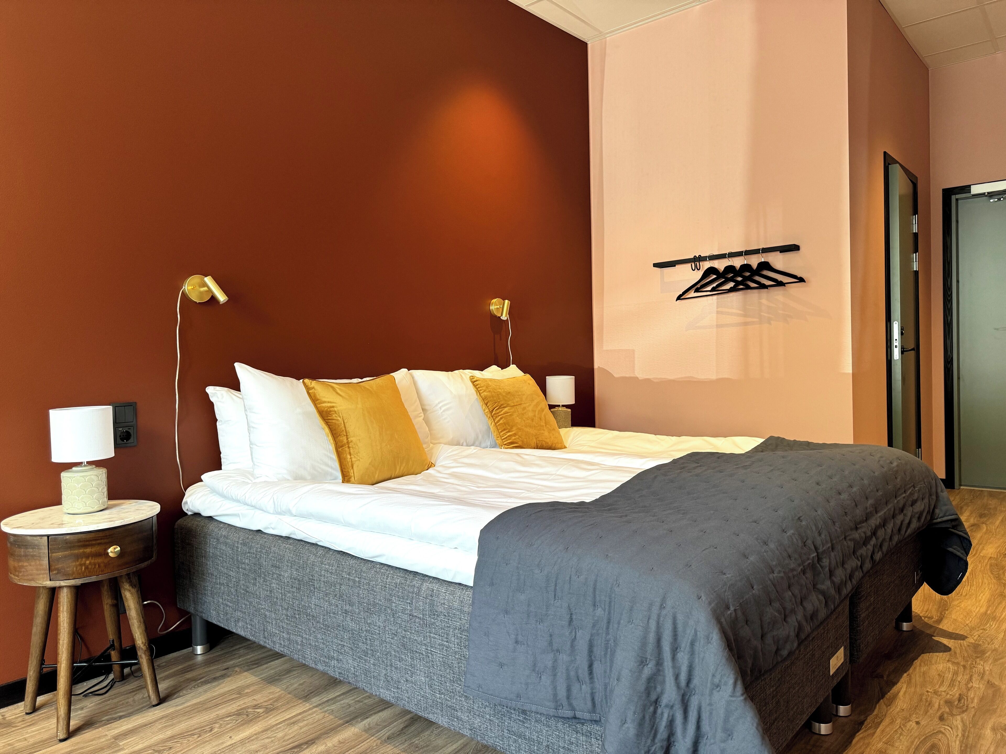 Quarto de hotel moderno com uma cama grande, mesa de cabeceira com abajur e cabideiro fixado na parede.
