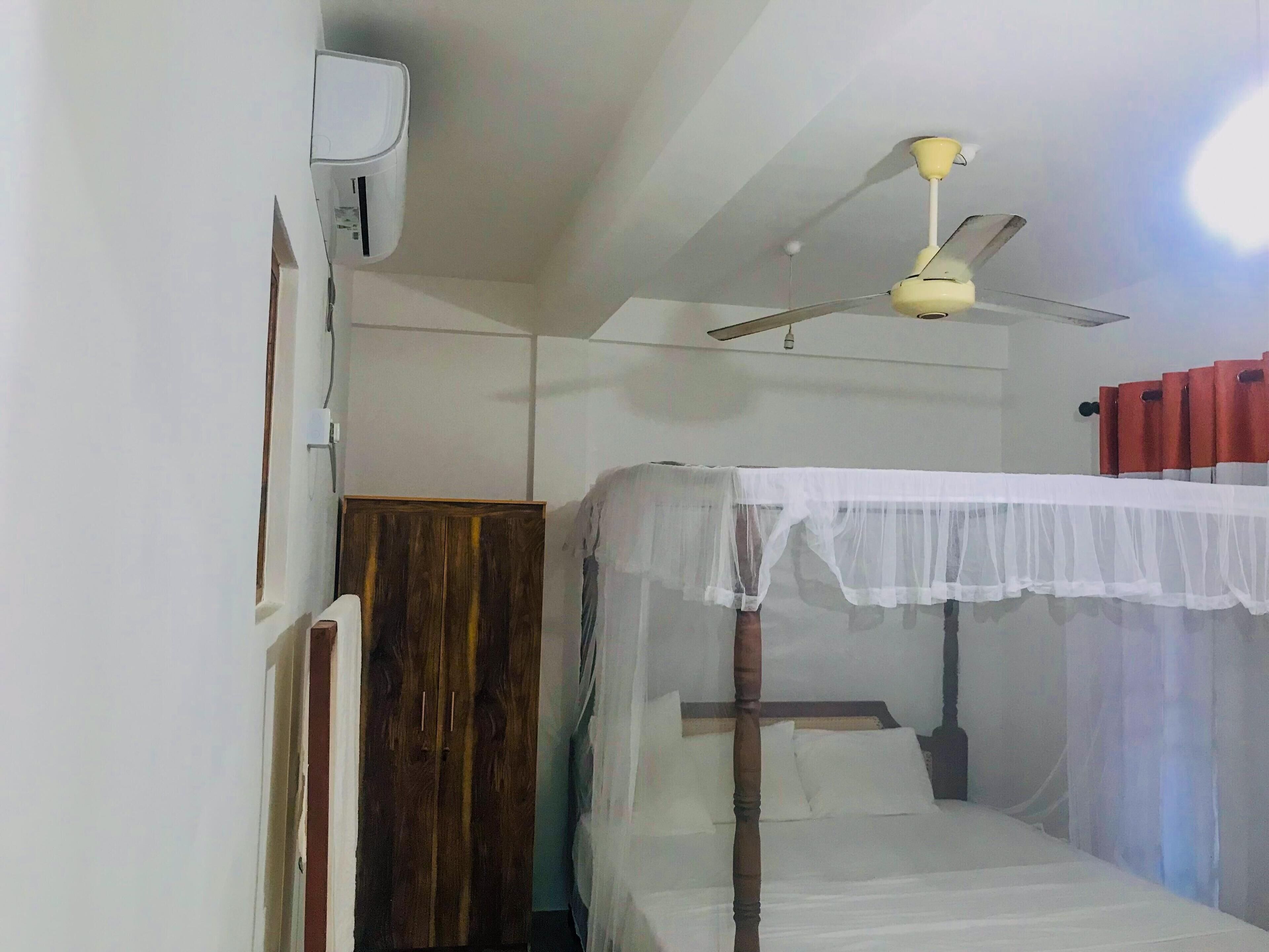 Uma cama de solteiro com mosquiteiro, um guarda-roupa de madeira, um ventilador de teto e um ar condicionado.