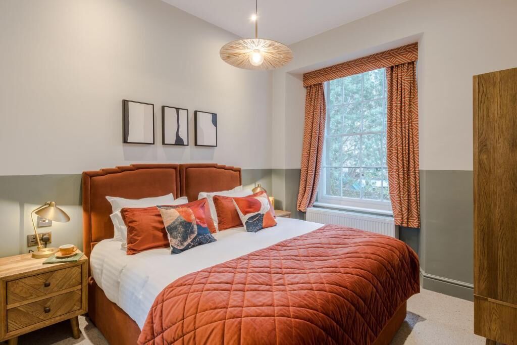 Quarto com uma cama grande, mesa de cabeceira com abajur, janela com cortinas e quadros nas paredes.