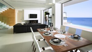 Uma sala de jantar moderna com uma mesa de madeira posta para refeição, um sofá preto e uma TV fixada na parede.