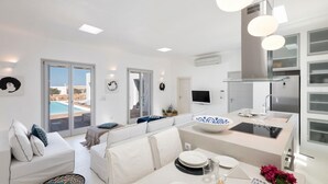 Uma sala de estar moderna e bem iluminada, com sofá, mesa de jantar e área de cozinha. É visível uma piscina através das portas de vidro.