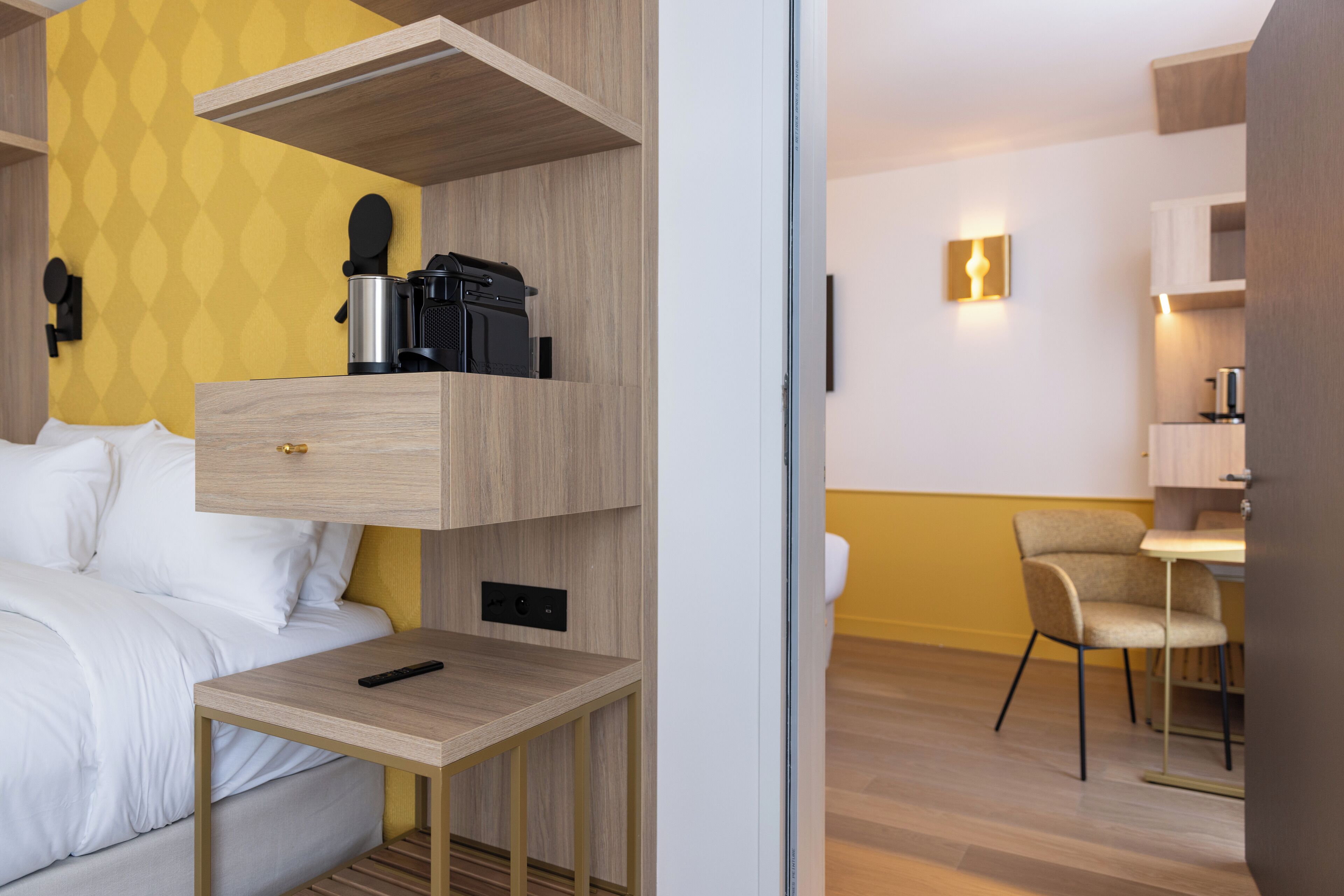 Quarto de hotel moderno com cama, uma mesinha, escrivaninha e uma cadeira. Há uma máquina de café na escrivaninha e uma prateleira fixada na parede acima da cama.