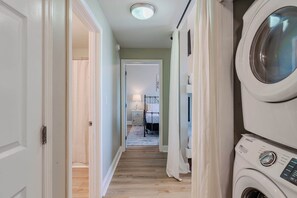 Área de lavanderia com máquina de lavar e secadora empilhadas, corredor que leva a um quarto com cama e mesa de cabeceira, e banheiro com cortina de chuveiro.