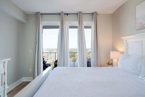 Quarto com cama grande, vista para uma varanda e mesa de cabeceira branca com abajur.