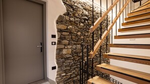 Uma escadaria moderna com degraus de madeira e guarda-corpos de metal, que leva a um espaço com parede de pedra e uma porta fechada.