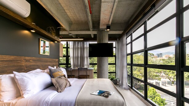 Quarto de hotel moderno com uma cama grande, televisão de tela plana e vista da cidade por grandes janelas.