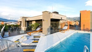 Uma área de piscina no terraço com espreguiçadeiras, um pequeno edifício e uma vista ampla da cidade e das montanhas ao fundo.