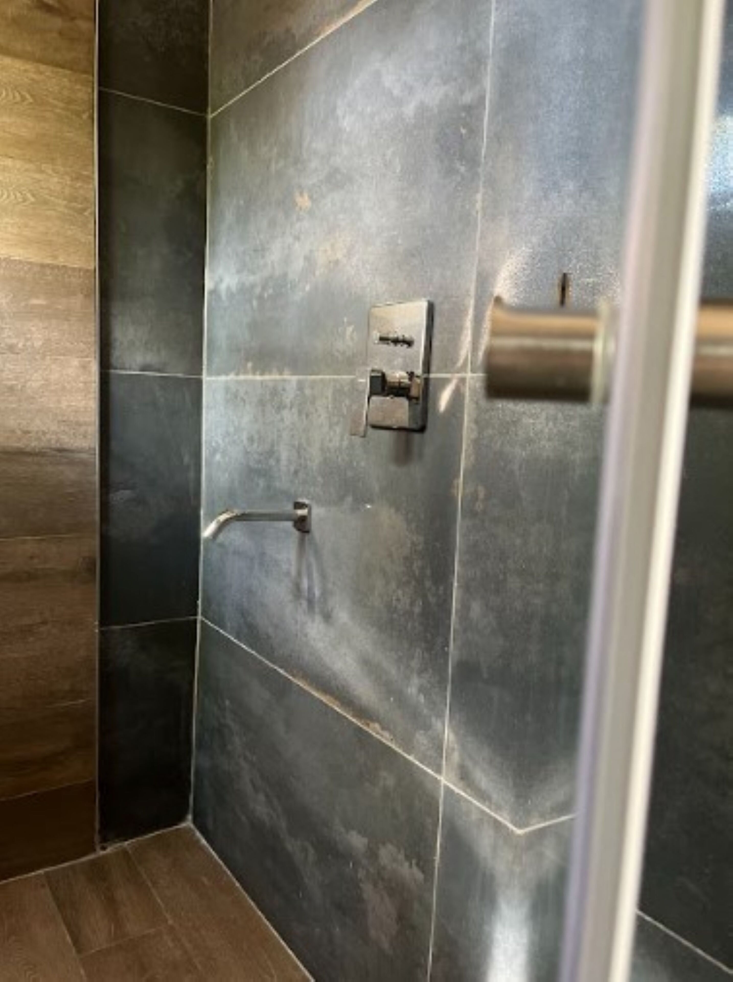 Um banheiro com azulejos cinza escuro, piso de madeira e chuveiro com maçaneta metálica.