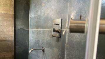 Um banheiro com azulejos cinza escuro, piso de madeira e chuveiro com maçaneta metálica.