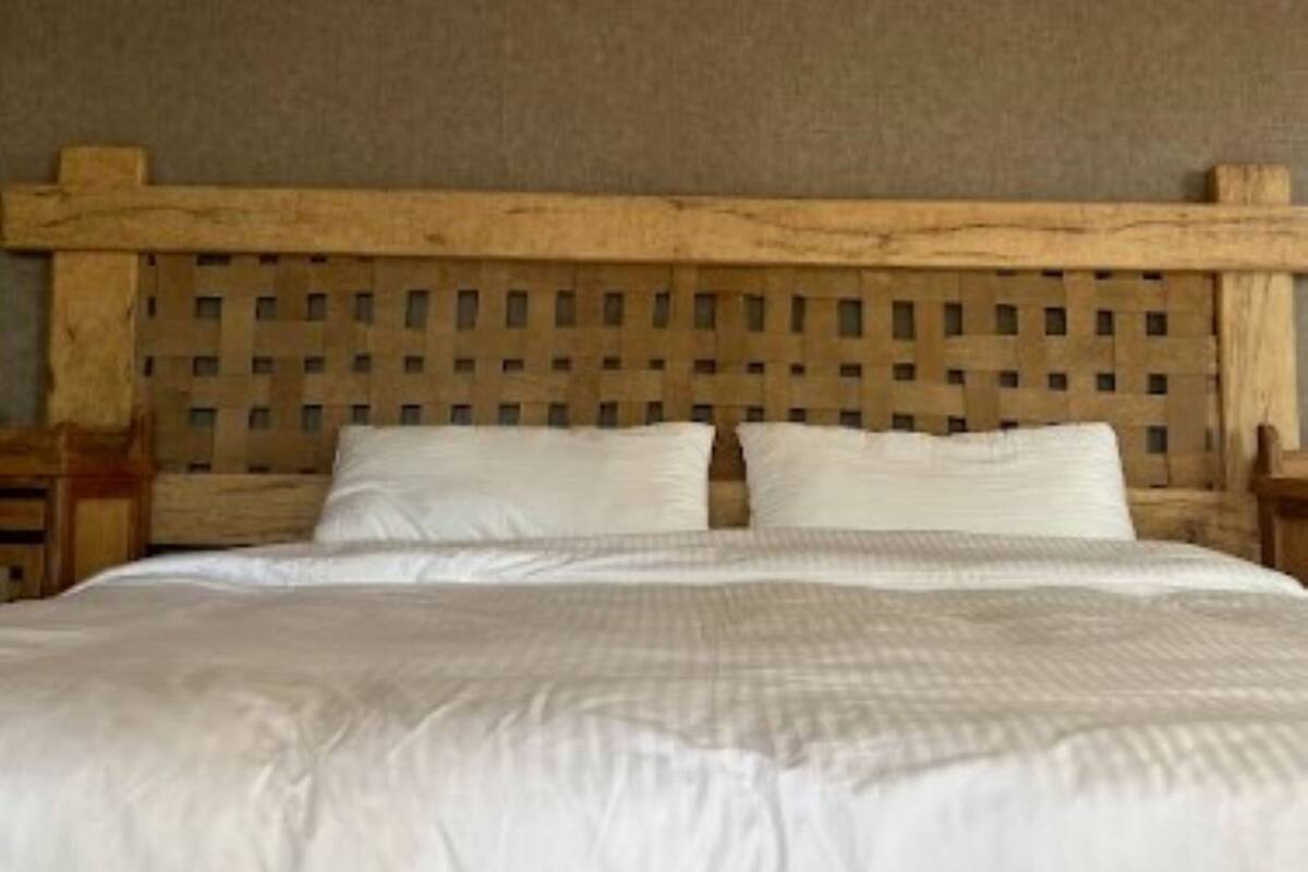 Cama com cabeceira de madeira, roupa de cama branca e parede lisa ao fundo.
