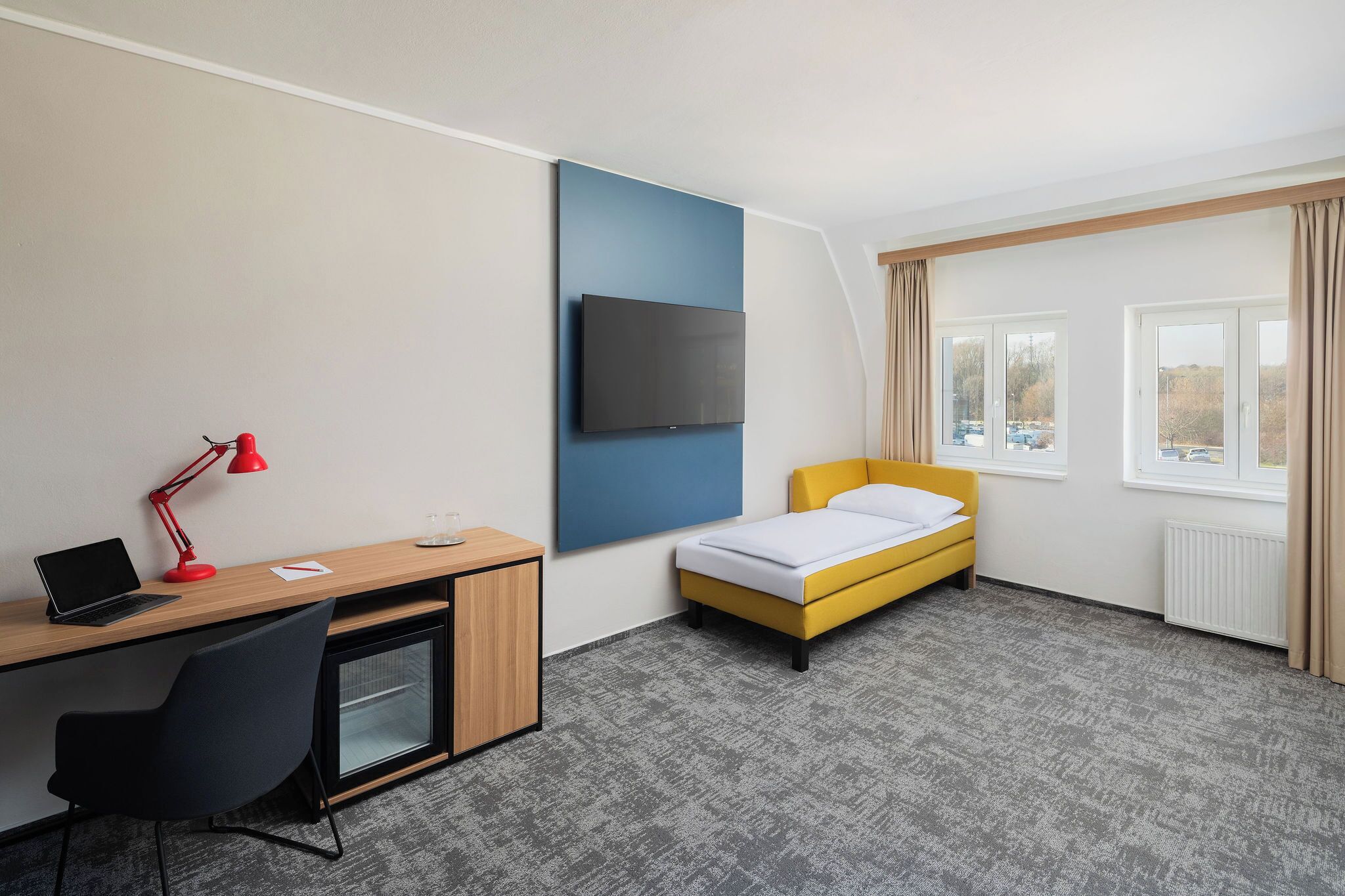 Quarto de hotel com cama, escrivaninha com laptop, televisão na parede e janela com cortinas.