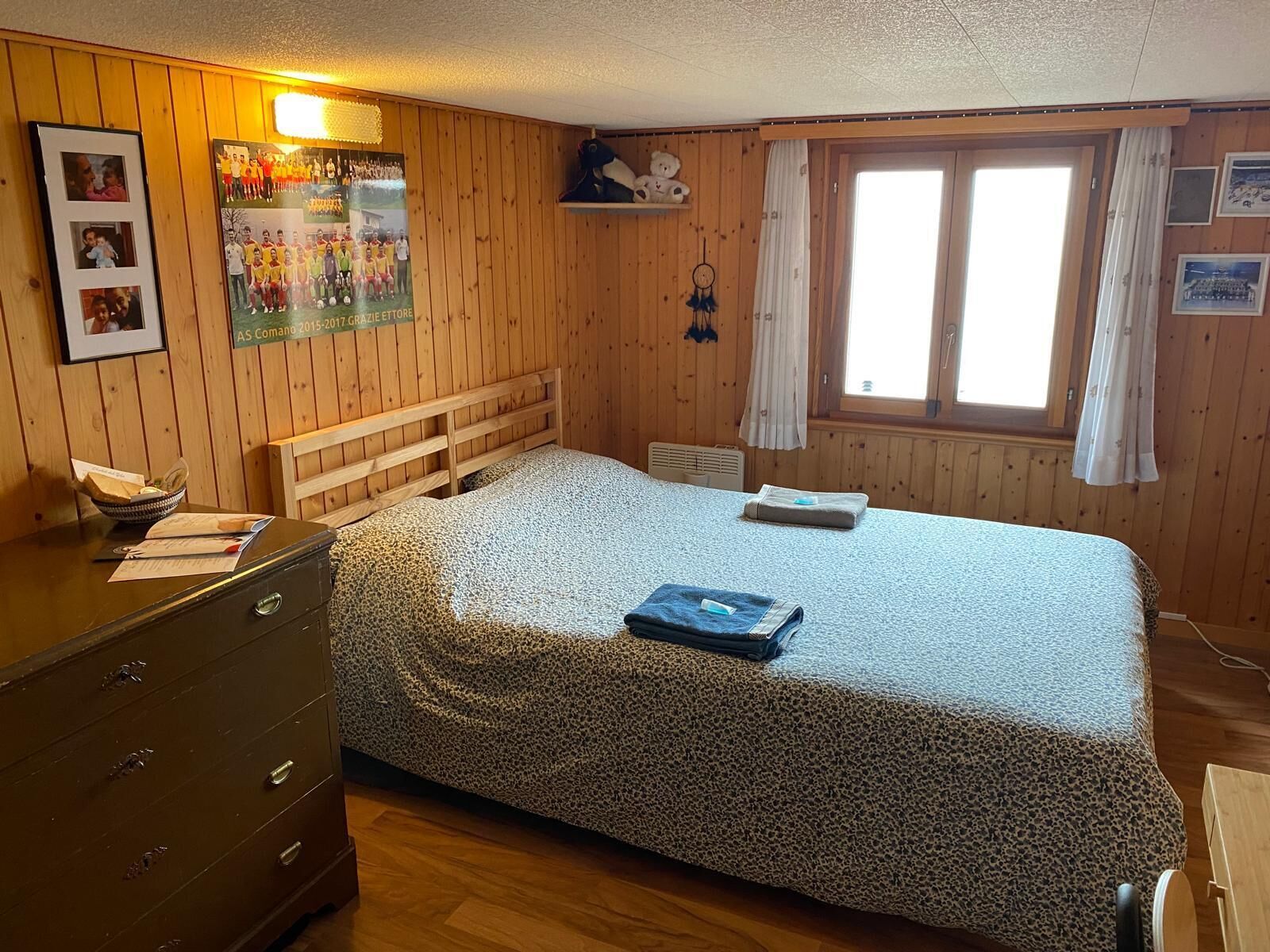 Um quarto com paredes de madeira, uma cama, uma cômoda, um cartaz e uma janela com cortinas.