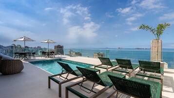 Área de piscina no terraço com espreguiçadeiras e guarda-sóis, com vista para a paisagem urbana e o mar.
