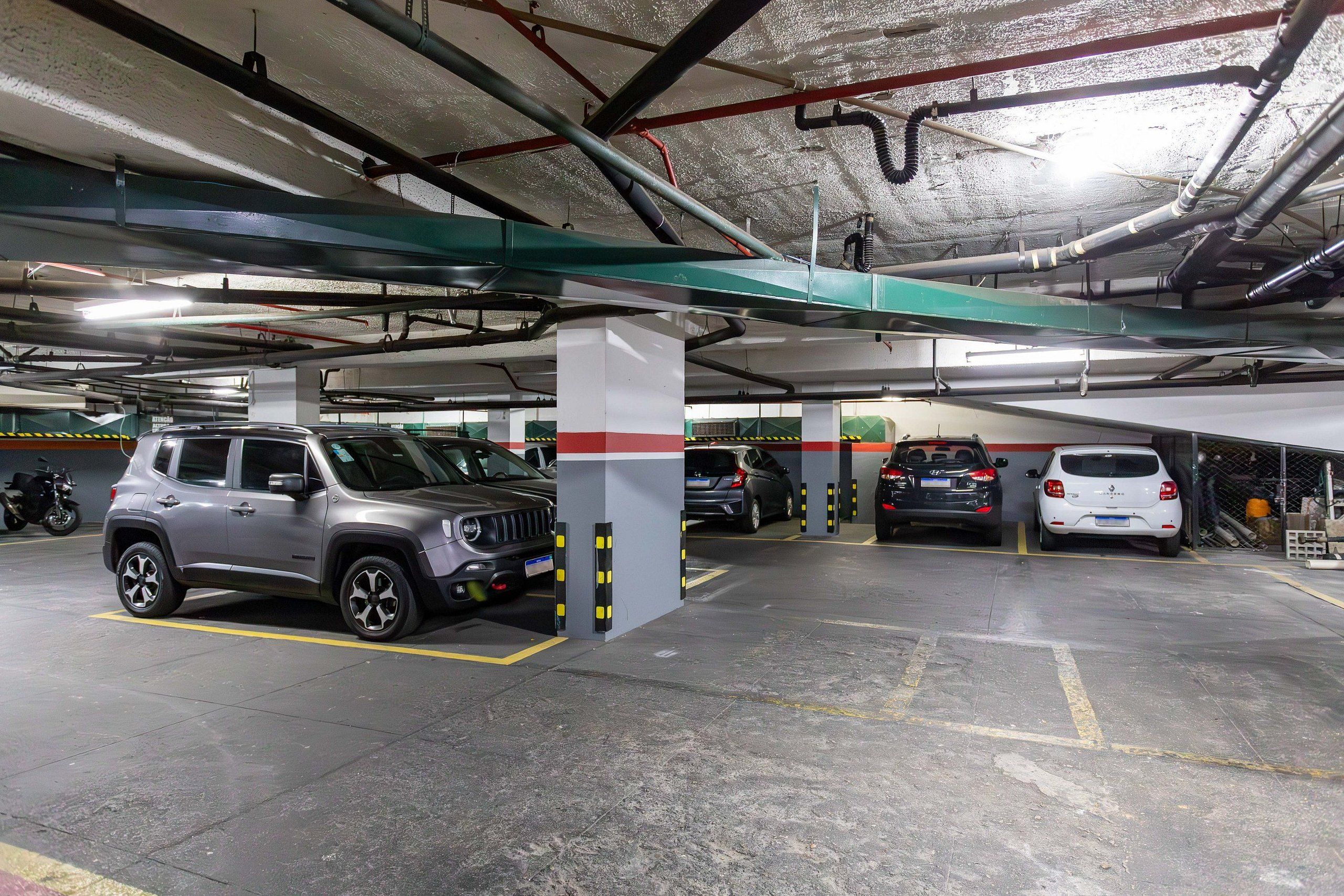 Garagem subterrânea com vários níveis, vários carros estacionados, pilares de concreto e tubulações no teto.