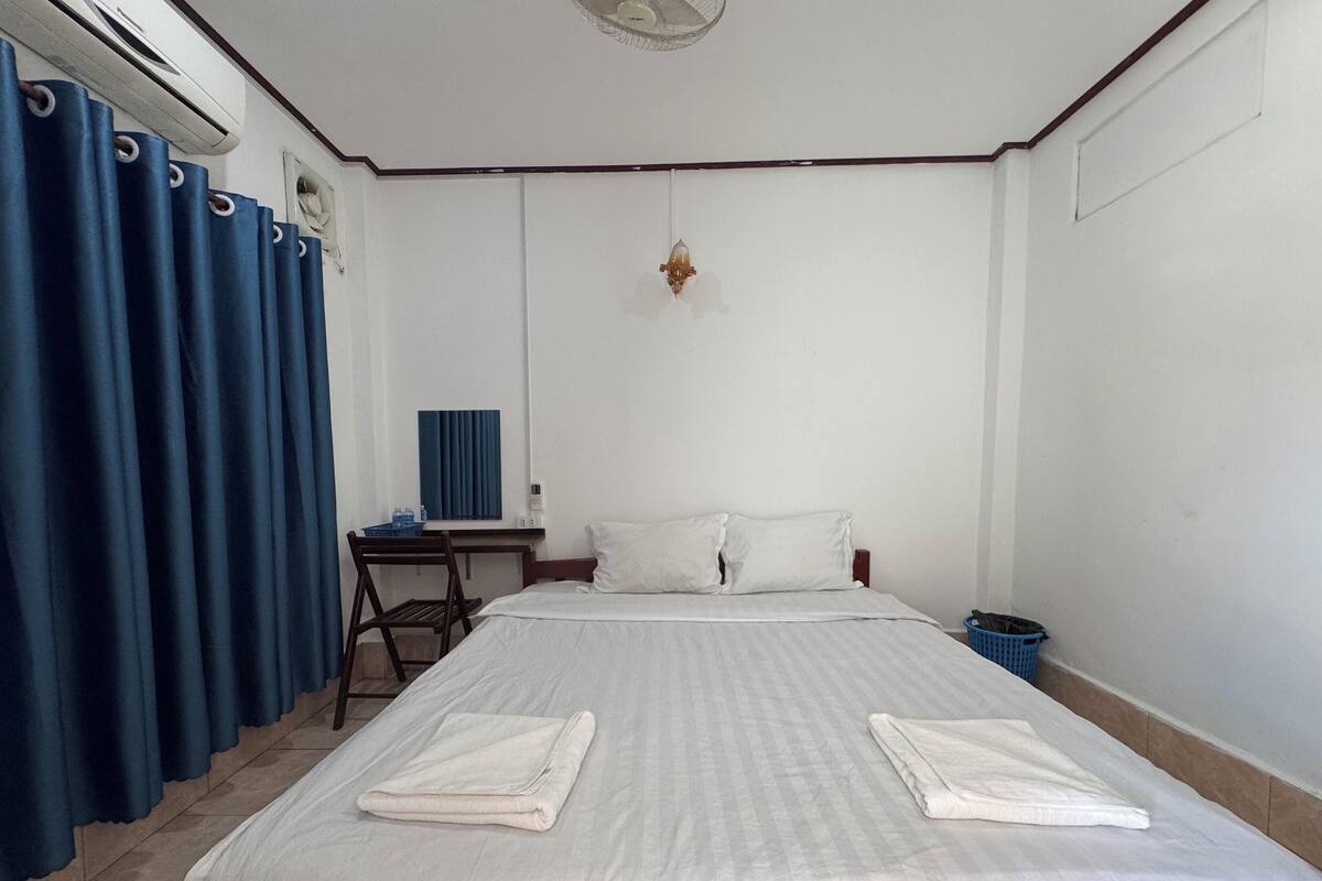 Quarto de hotel com cama, escrivaninha, cadeira, cortina azul e luminária fixada na parede.