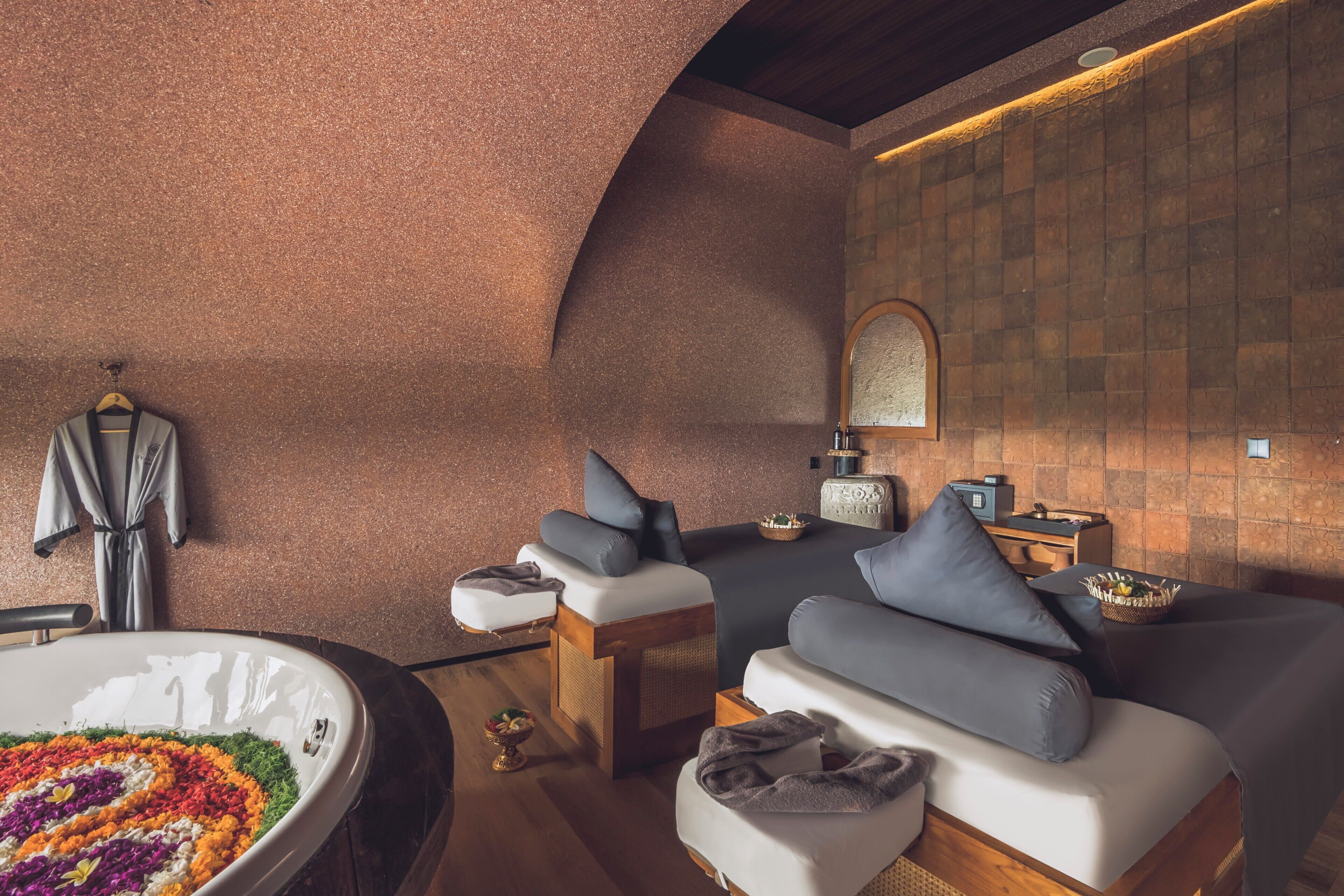 Sala de spa com mesa de massagem, duas cadeiras de massagem, um banco de madeira, um arranjo floral decorativo e um espelho fixado na parede.