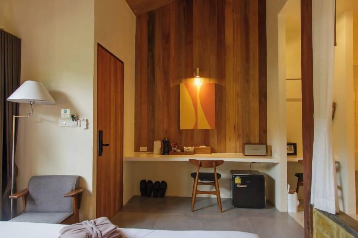 Quarto de hotel com uma cama, uma escrivaninha com cadeira, uma luminária, um cesto de lixo pequeno e um painel de parede de madeira.