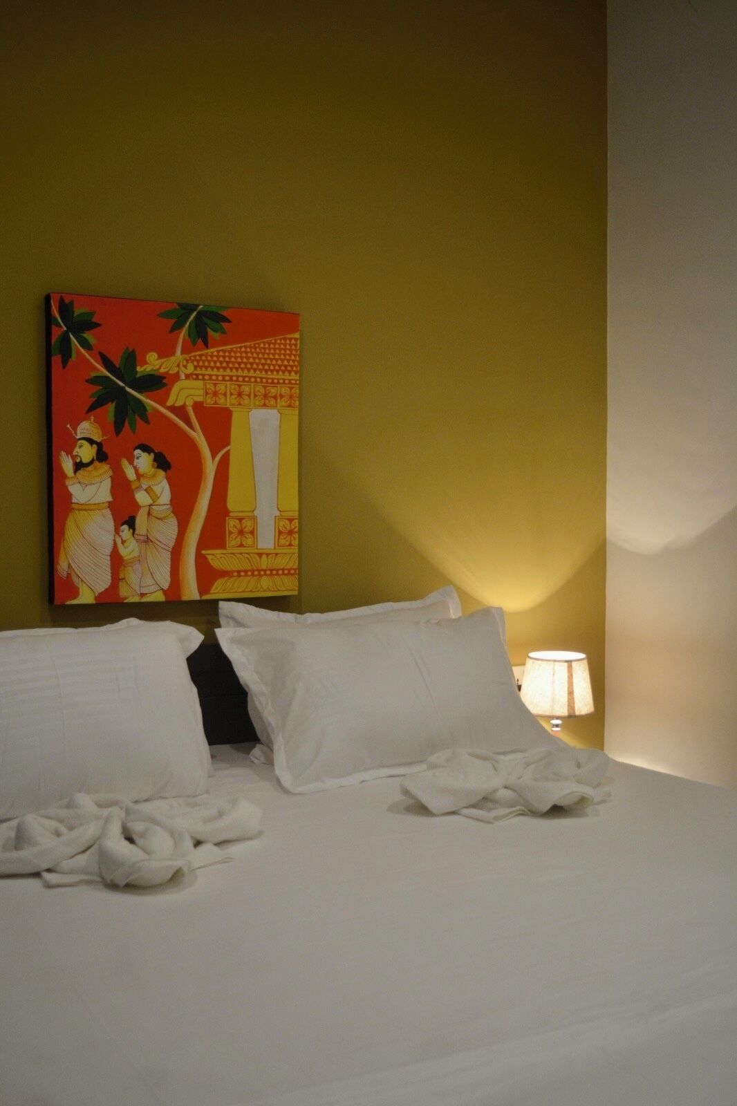 Quarto de hotel com cama, almofadas brancas, abajur e um quadro na parede.