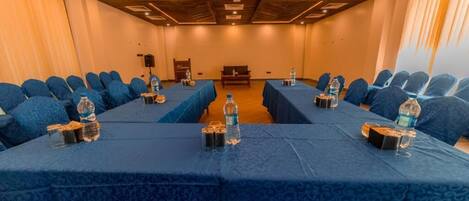 Sala de conferências com cadeiras estofadas em azul e mesas, equipada com garrafas de água e copos.