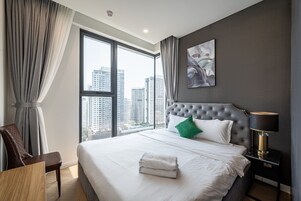 Quarto de hotel com uma grande janela, cama com roupa de cama branca, travesseiro verde e cabeceira cinza.