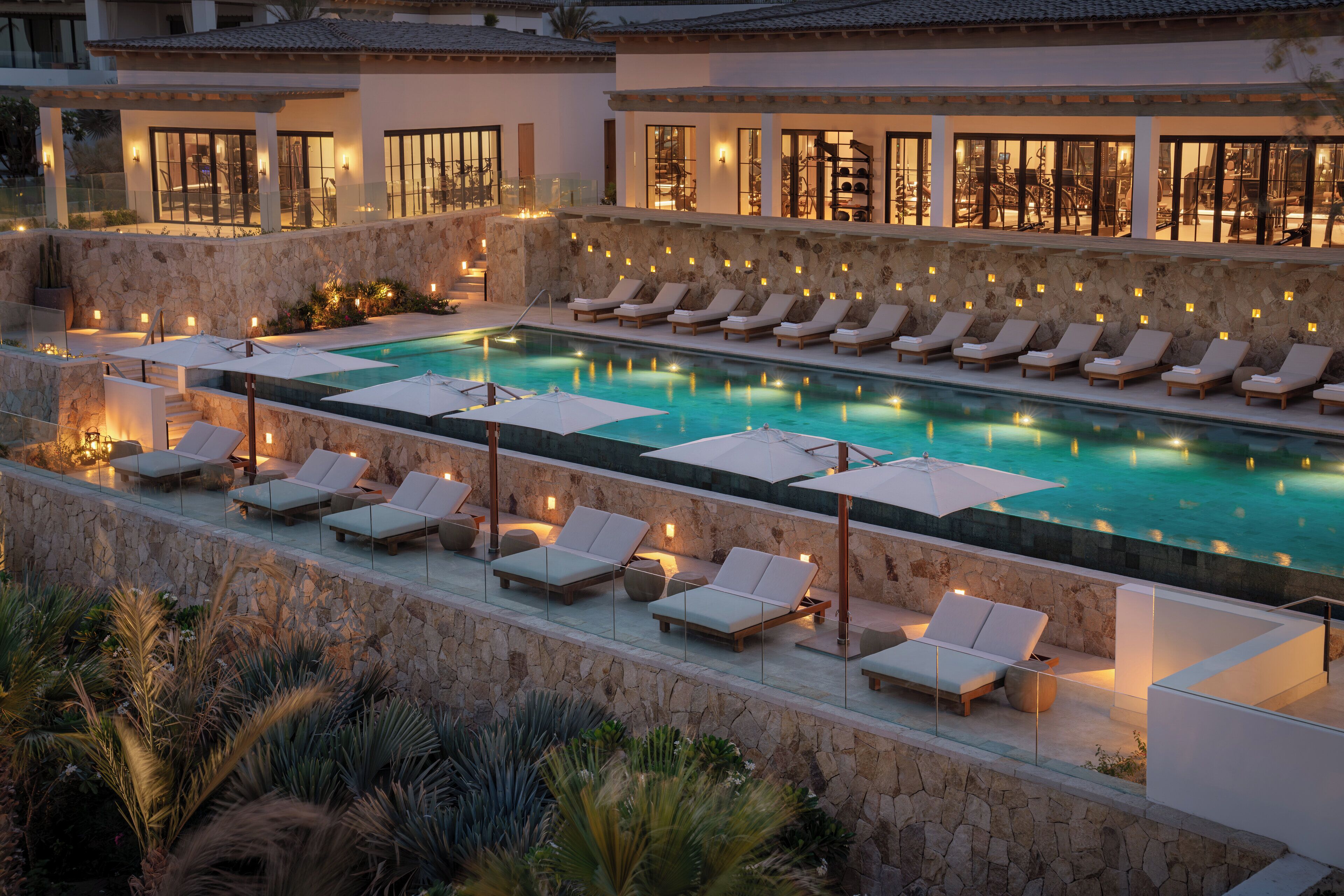 Uma área de piscina com espreguiçadeiras e guarda-sóis, cercada por paredes de pedra e um edifício com grandes janelas.