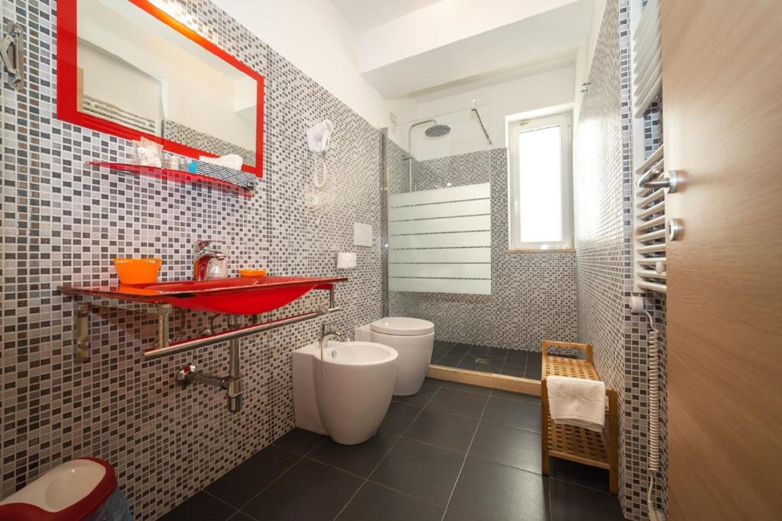 Casa de banho moderna com painel de azulejos, espelho com moldura vermelha, lavatório branco, duas sanitas e área de chuveiro.