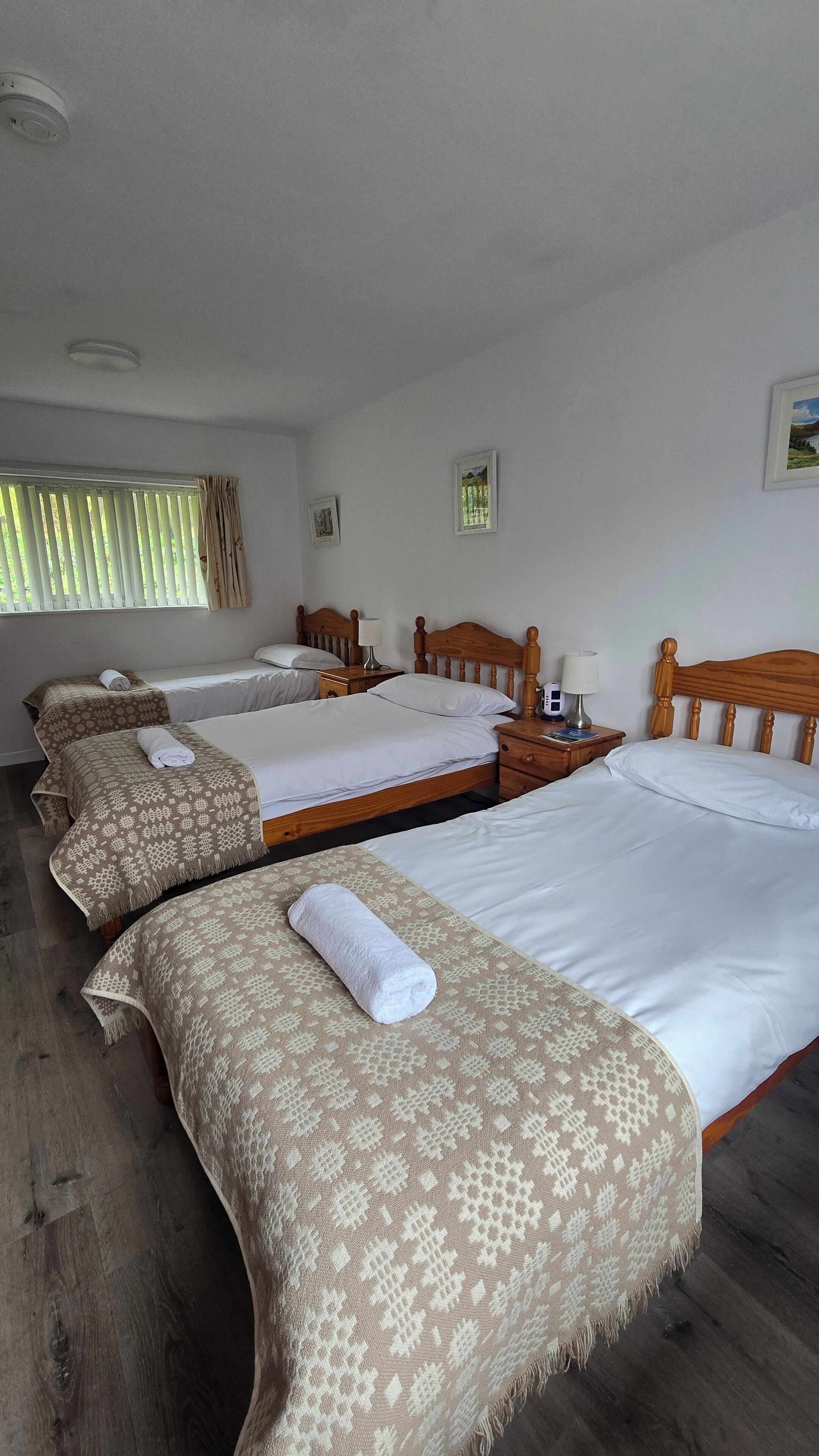 Quarto de hotel com quatro camas, cada uma com cabeceira de madeira, roupa de cama branca e manta decorativa. Há mesas de cabeceira com luminárias e uma janela com persianas.