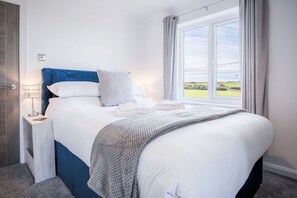 Cama bem arrumada com roupa de cama branca e azul, um abajur e vista para uma paisagem verde através de uma janela.
