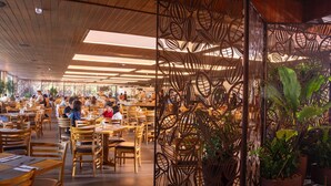 Um restaurante espaçoso com mesas e cadeiras de madeira, divisória metálica decorativa e plantas em vasos.