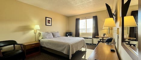 Quarto de hotel com cama, escrivaninha com telefone, cadeira, abajur, espelho e janela com cortinas.