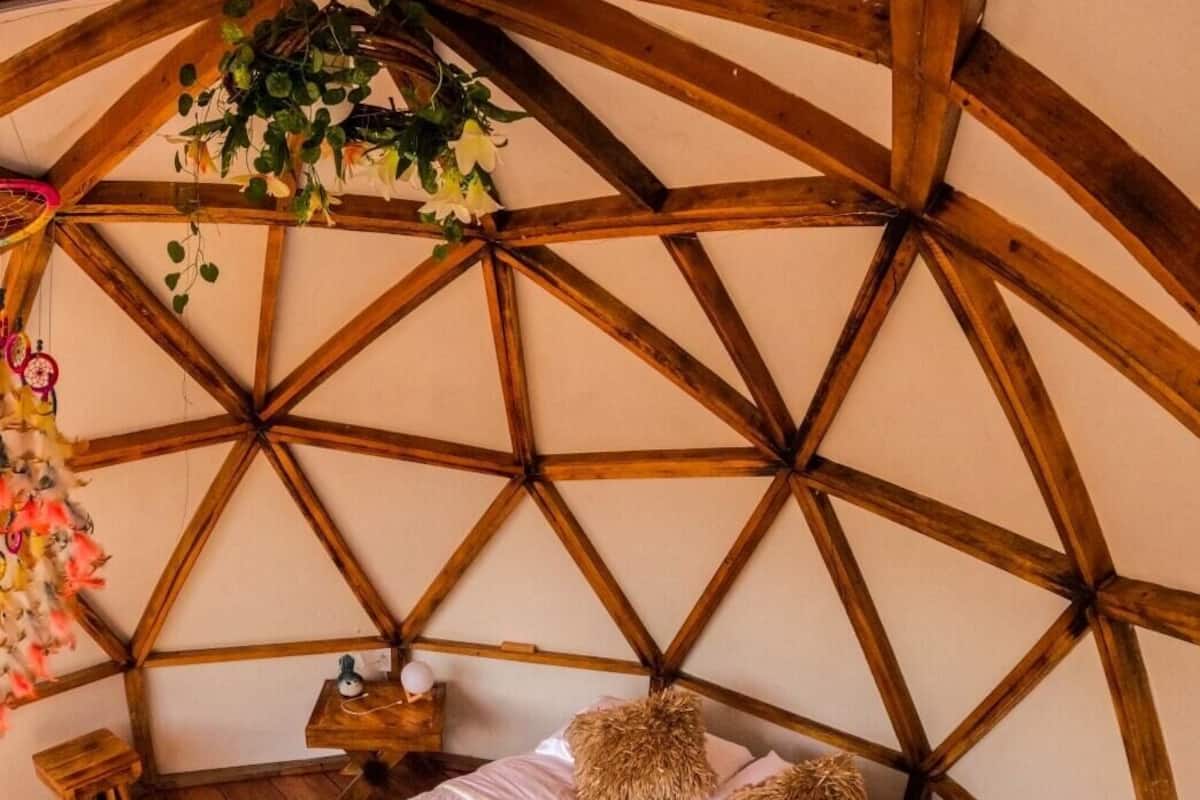 Um quarto com teto de madeira geométrico, uma cama com cobertor branco e uma pequena mesa de madeira com abajur.