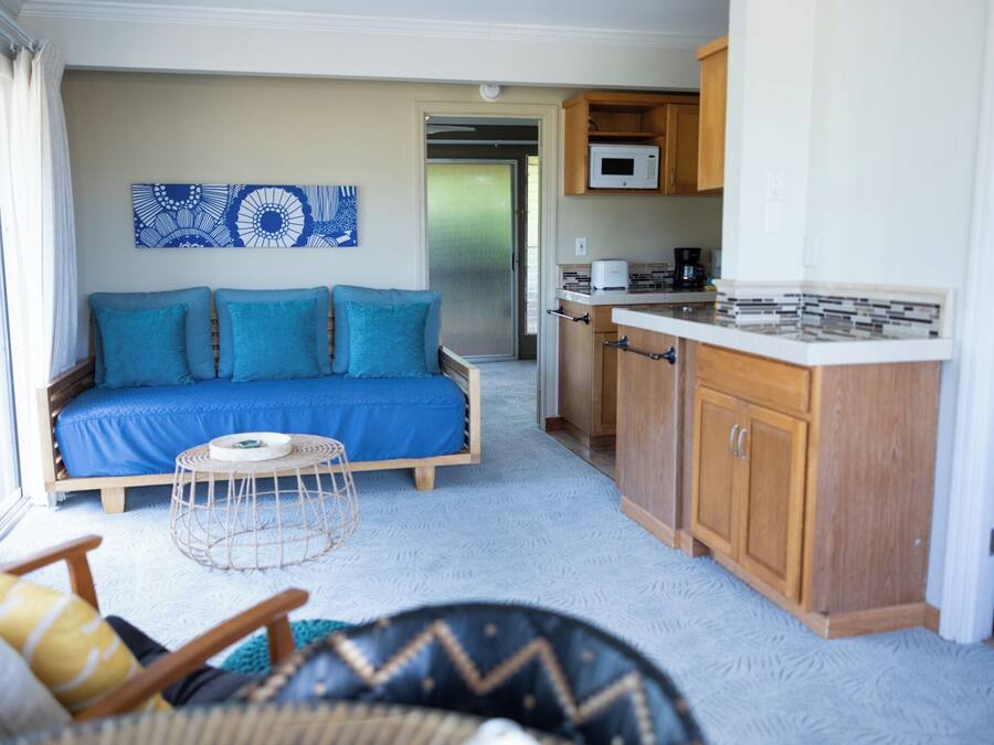 Uma sala de estar com um sofá azul, uma mesa redonda pequena e um espaço de cozinha com armários de madeira e micro-ondas.
