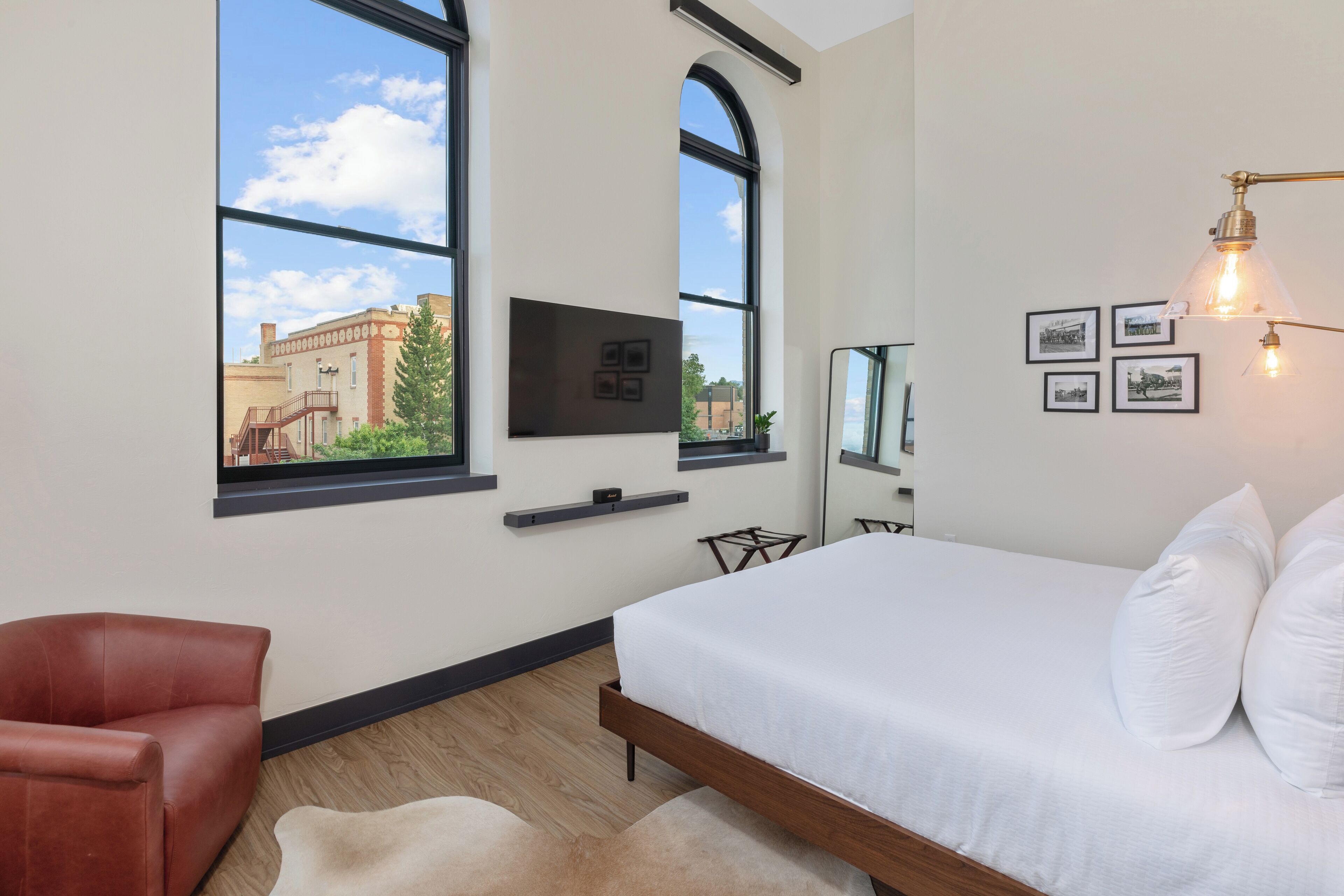 Quarto com cama grande, uma cadeira vermelha, piso de madeira, TV na parede e vista para a cidade pelas janelas.