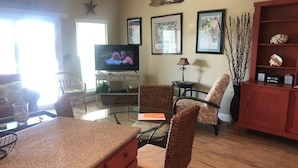 Uma sala de estar com televisão, cadeiras de vime, mesa de centro de vidro, um armário vermelho e quadros nas paredes.