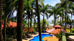 Área da piscina com espreguiçadeiras e guarda-sóis, cercada por palmeiras e vista para a praia.