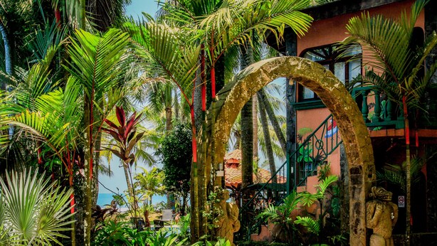 Um jardim exuberante com plantas tropicais, um arco de pedra e um edifício com fachada vermelha e detalhes verdes.