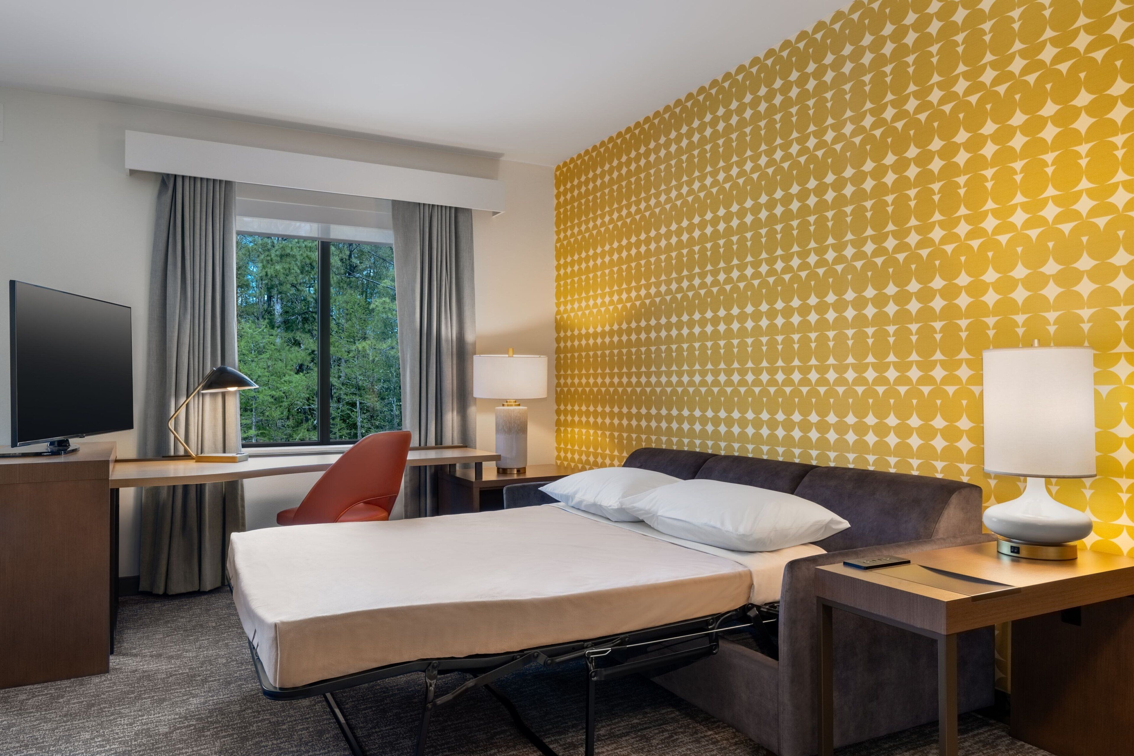 Quarto de hotel com cama, escrivaninha, cadeira, televisão e janela com cortinas.