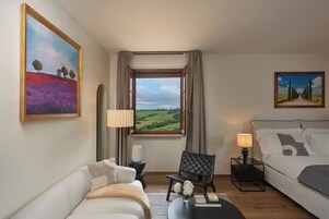 Quarto de hotel com uma cama, um sofá, uma cadeira, uma mesinha com abajur e uma janela com vista para uma paisagem verde.