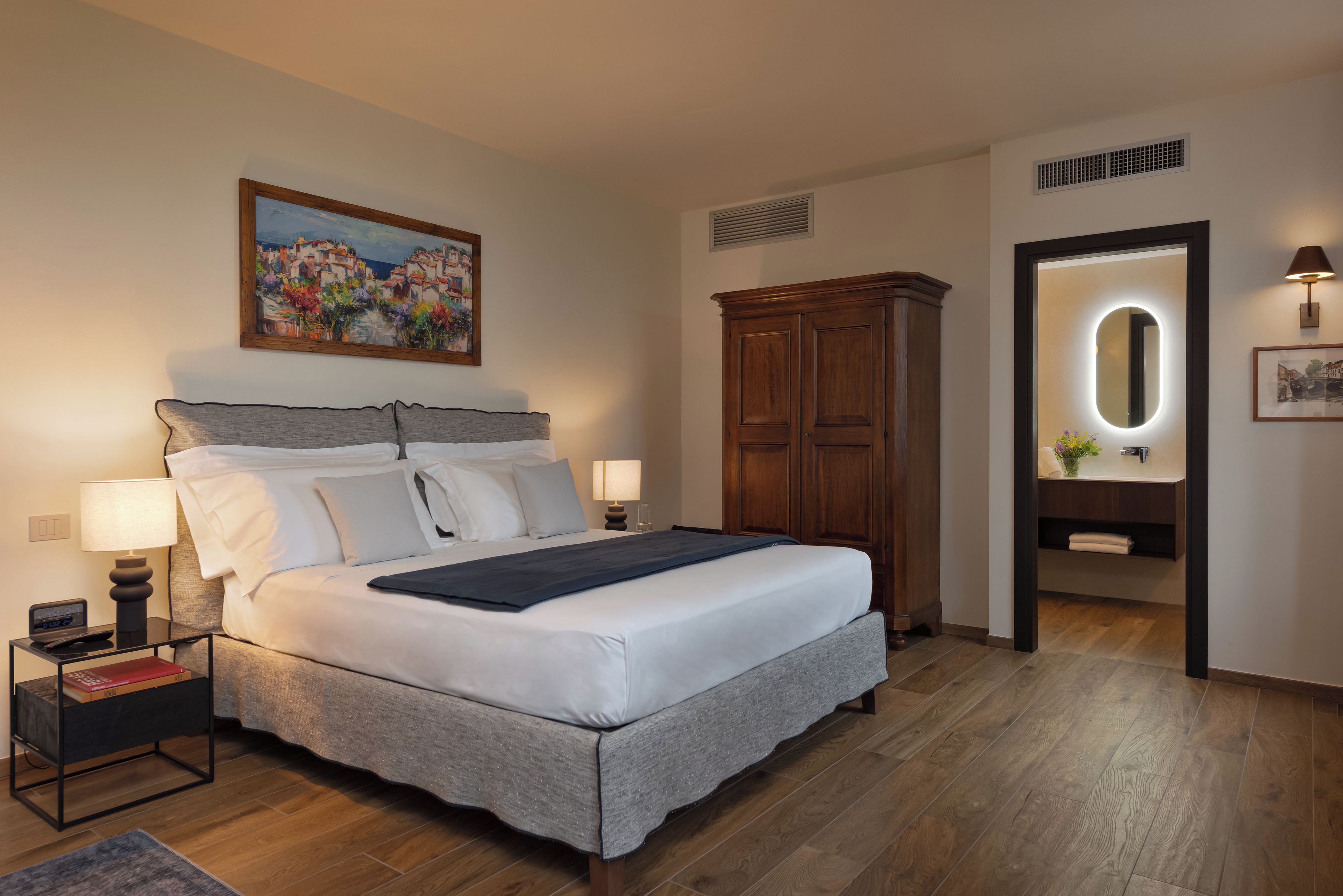 Quarto bem arrumado com cama, mesas de cabeceira, guarda-roupa, espelho e um quadro na parede.