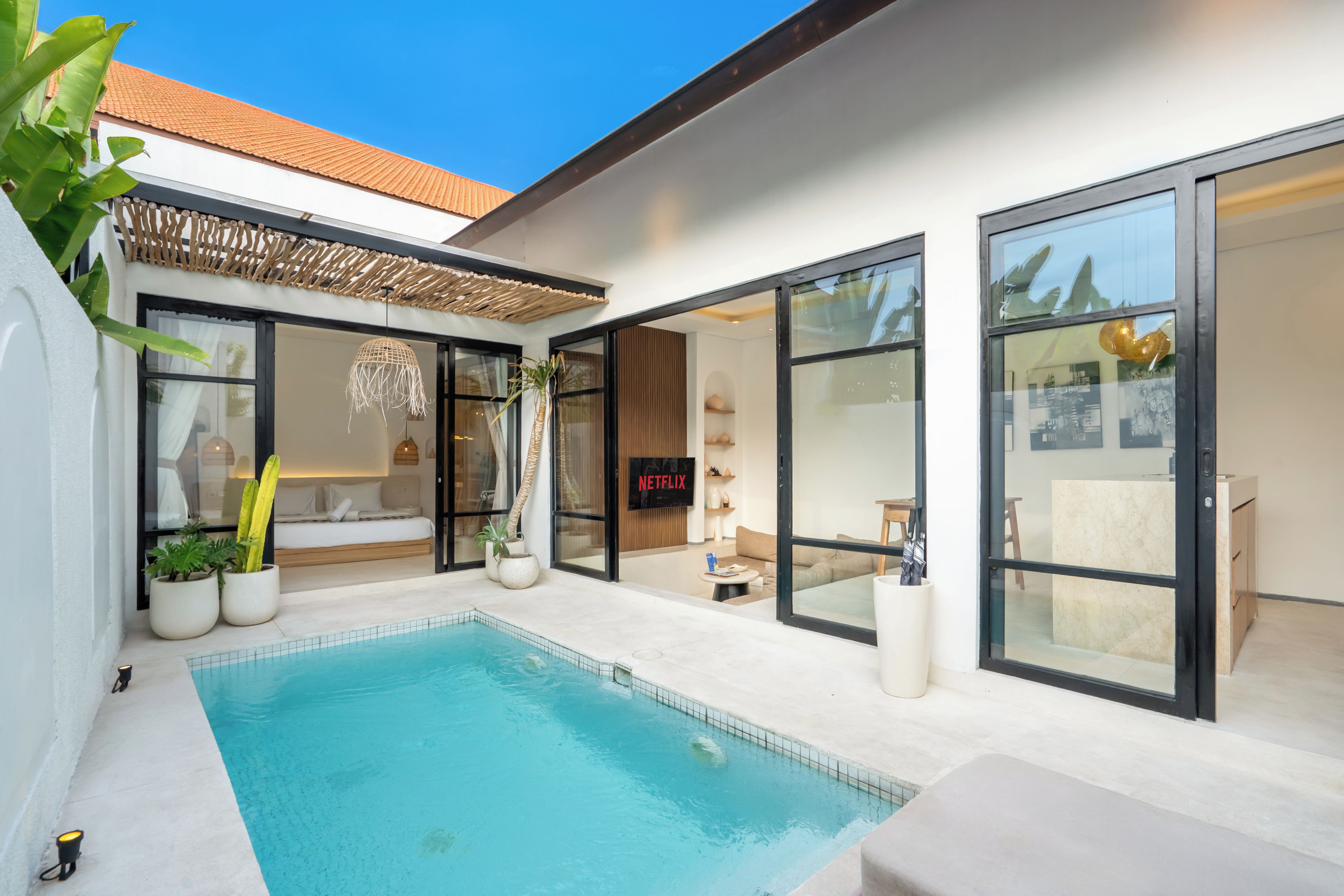 Uma área moderna de piscina ao ar livre, com piscina azul cristalina, zona coberta com assentos e um grande painel de vidro deslizante que dá para uma sala de estar.